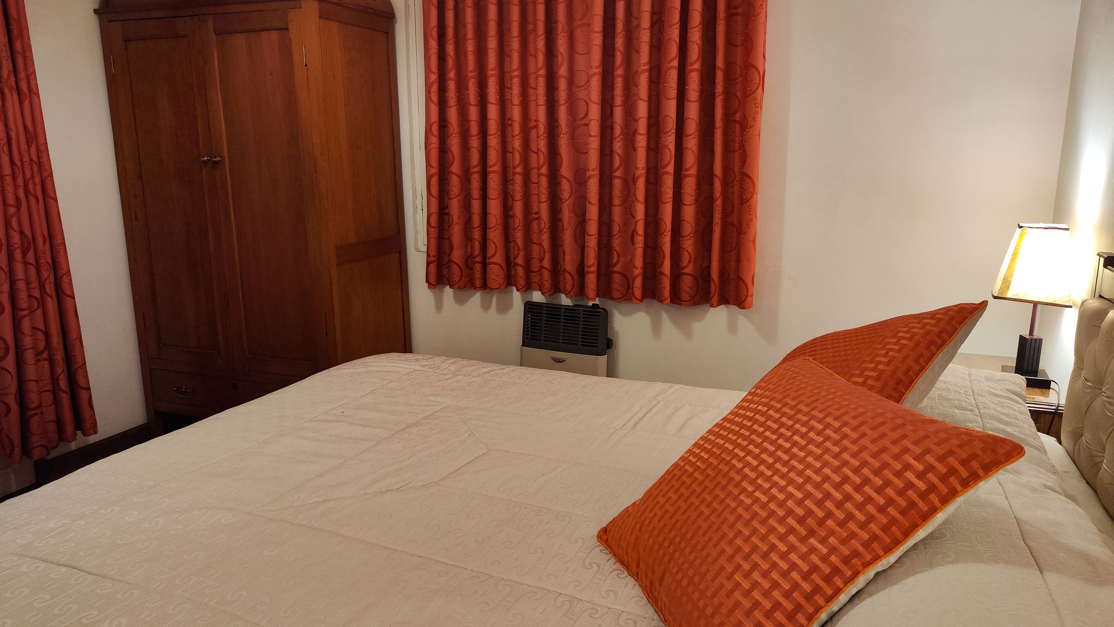 Quarto de hotel com cama, aquecedor, mesa de cabeceira com abajur e cortinas vermelhas.