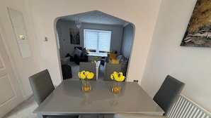 Uma sala de jantar com uma mesa cinza e quatro cadeiras, dois vasos com flores amarelas e vista para uma sala de estar com um sofá e uma janela.