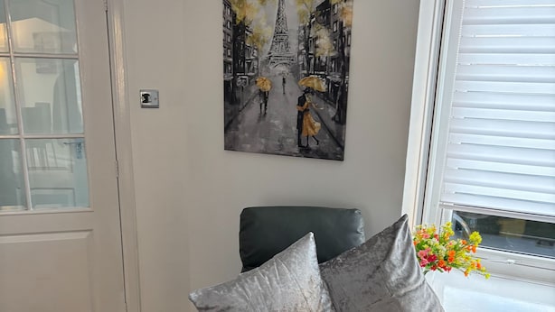 Quarto com sofá cinzento, pintura da Torre Eiffel, janela com persianas e radiador.
