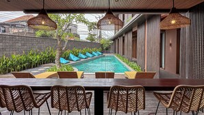 Uma área à beira da piscina com uma mesa comprida e cadeiras de vime, piscina com almofadas azuis e vista para a cidade.