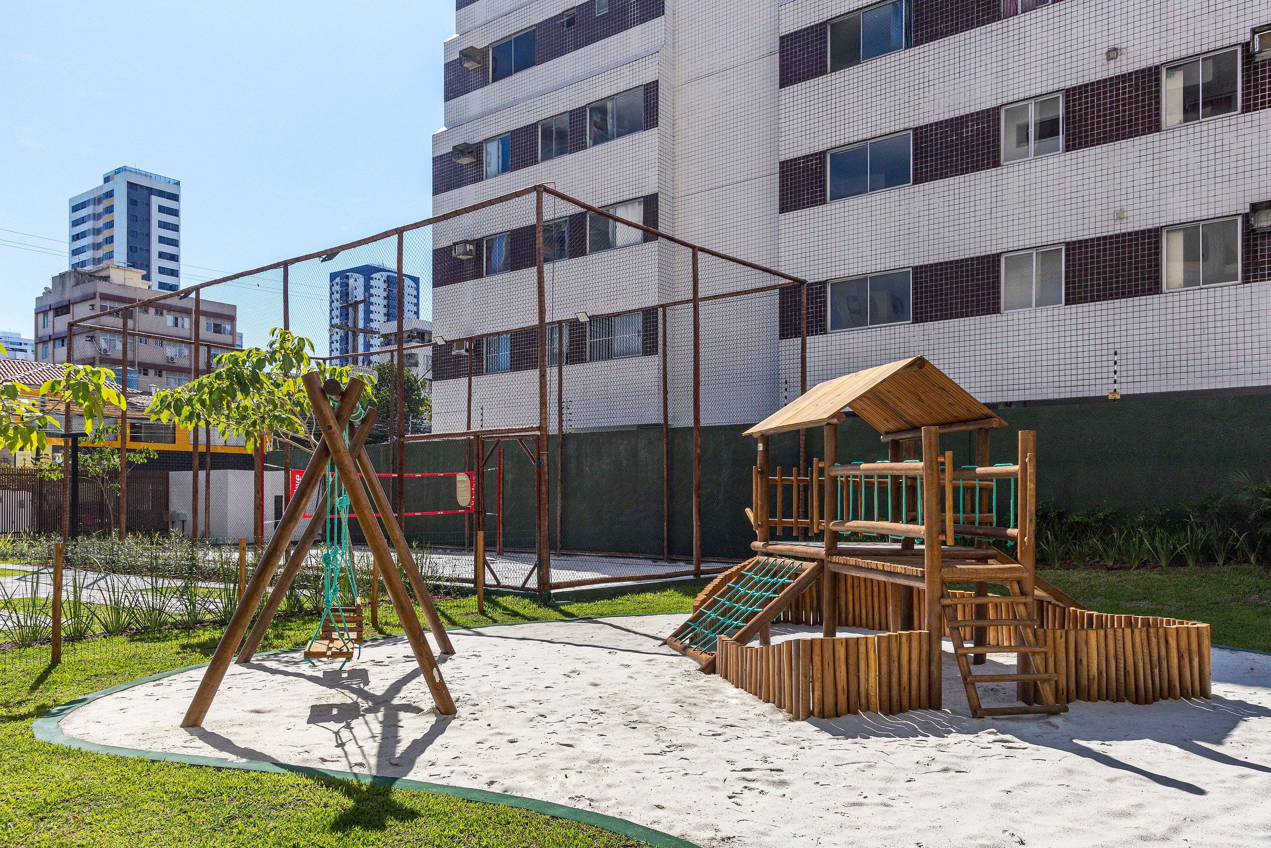 Um parquinho com balanços e uma estrutura de escalada, cercado por uma área de areia e um edifício ao fundo.