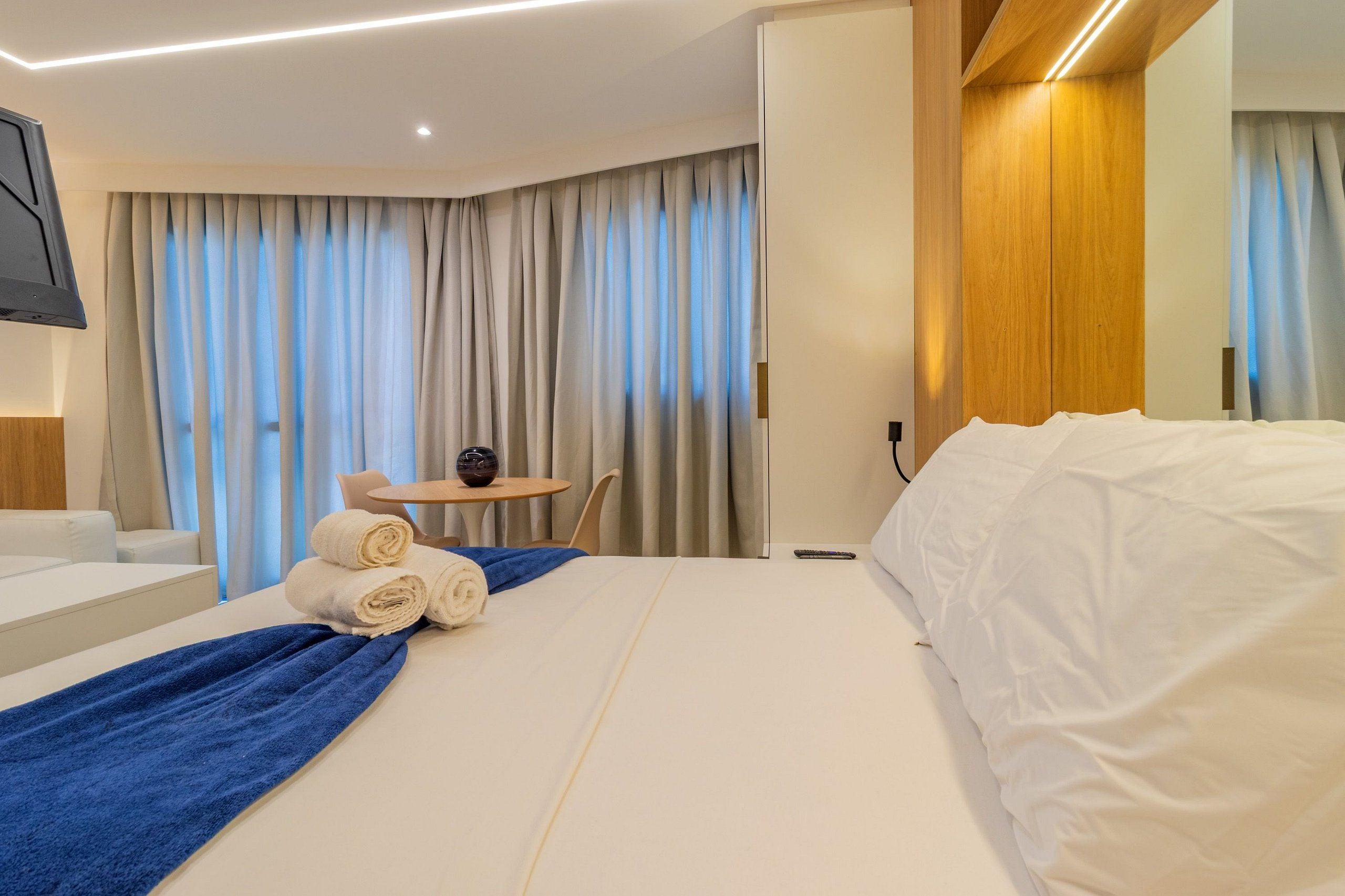Quarto de hotel moderno com uma cama bem arrumada, uma toalha azul e uma pequena mesa redonda com um vaso.