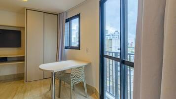 Quarto moderno com uma janela grande, uma secretária branca e uma cadeira.