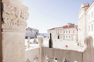 Varanda com uma garrafa de vinho e copos, com vista para uma praça de arquitetura clássica.