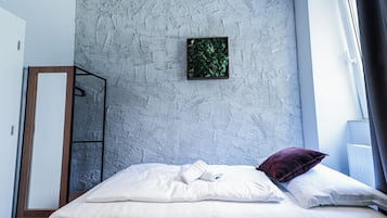 Quarto com cama, almofadas brancas, uma almofada roxa, planta emoldurada e guarda-roupa.