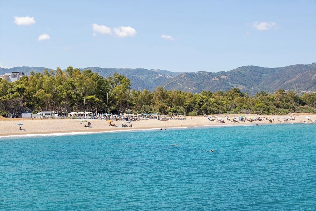 Uma praia com águas turquesa cristalinas, uma faixa de areia e colinas verdes ao fundo.