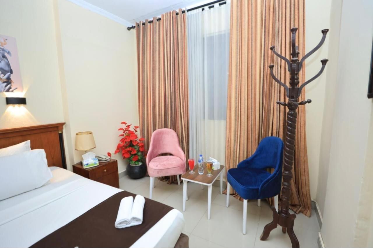 Quarto de hotel com cama, mesa de cabeceira, luminária, cadeira, mesa com bebidas, porta-casacos e cortinas.