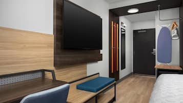 Quarto de hotel moderno com escrivaninha de madeira, um banco acolchoado azul, televisão de tela plana, guarda-roupa e uma cama com colcha estampada.