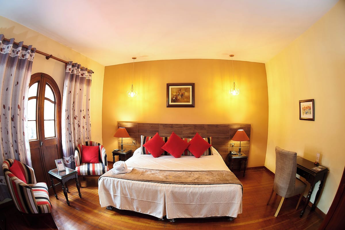 Quarto de hotel com uma cama grande, duas poltronas, uma escrivaninha e cabeceira de madeira.