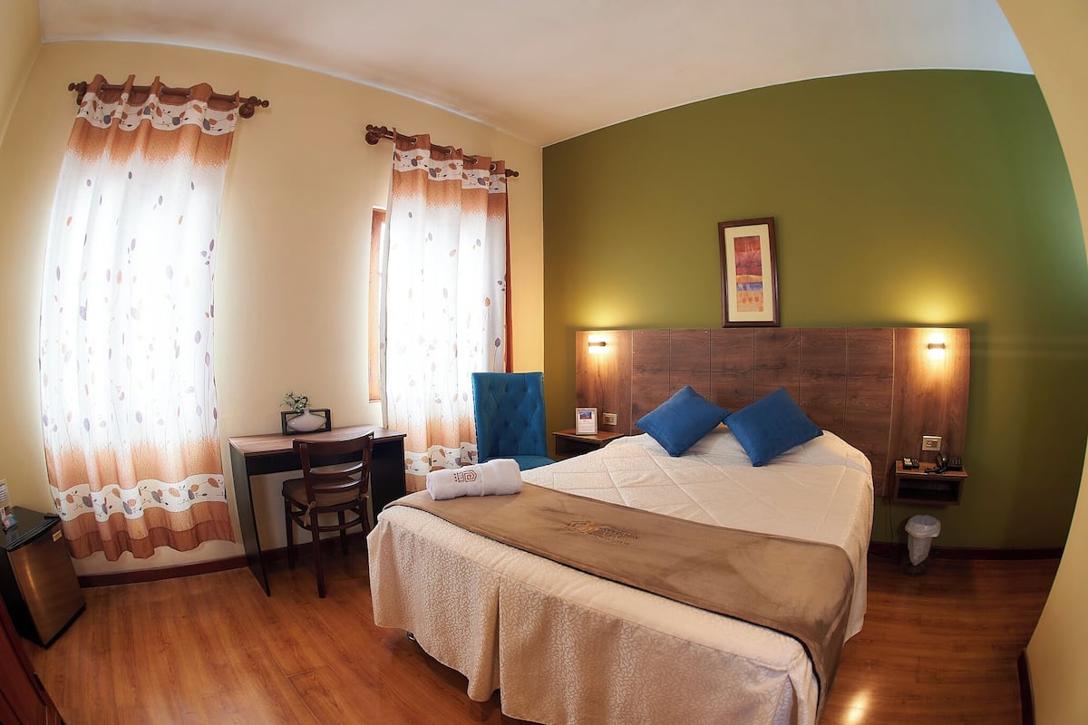 Quarto de hotel com uma cama grande, uma mesa com cadeira, uma mesinha e um cesto de lixo.