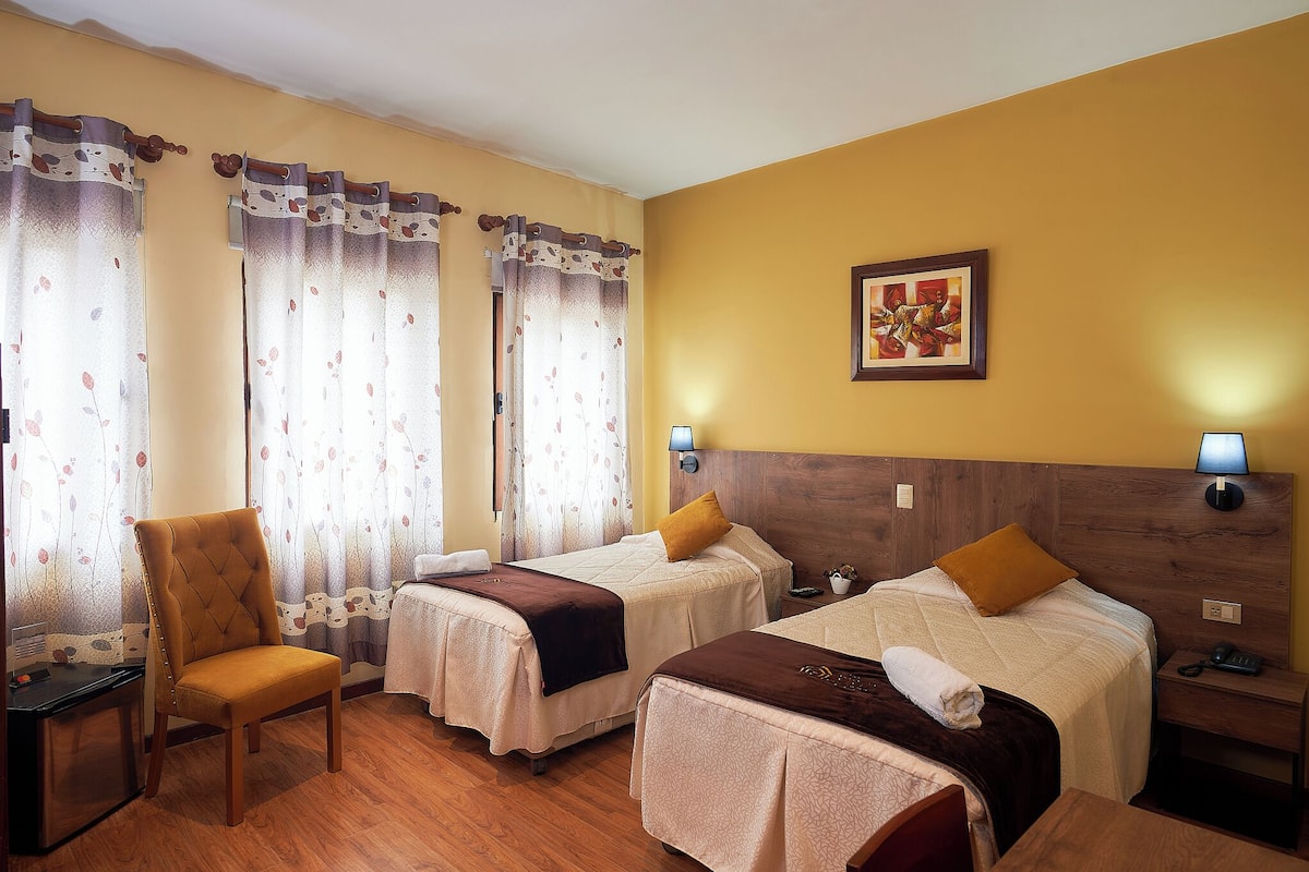 Quarto de hotel com duas camas, uma cadeira, uma escrivaninha e um abajur.
