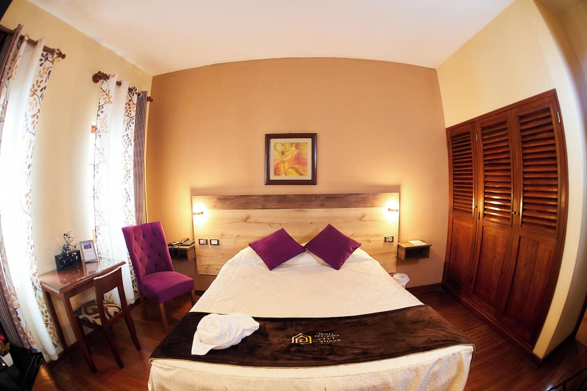 Quarto de hotel com cama, poltrona, escrivaninha, quadro na parede e guarda-roupa.