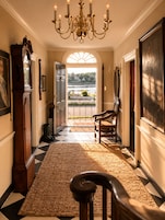 Um corredor com um candelabro, um banco de madeira, uma pintura grande e vista para um lago por uma porta em arco.