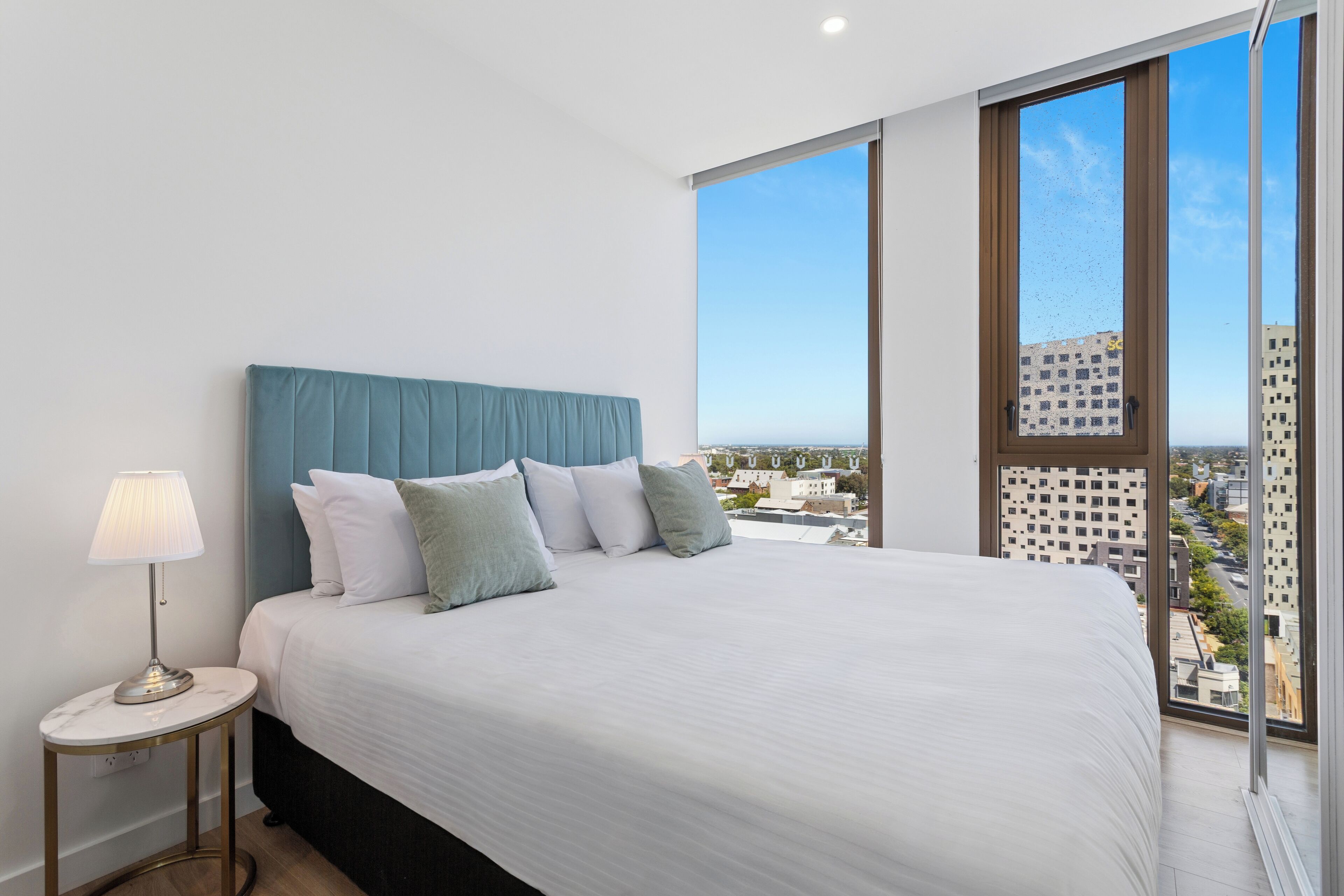 Quarto moderno com uma cama grande, uma mesa de cabeceira com abajur e vista para a cidade através de amplas janelas.