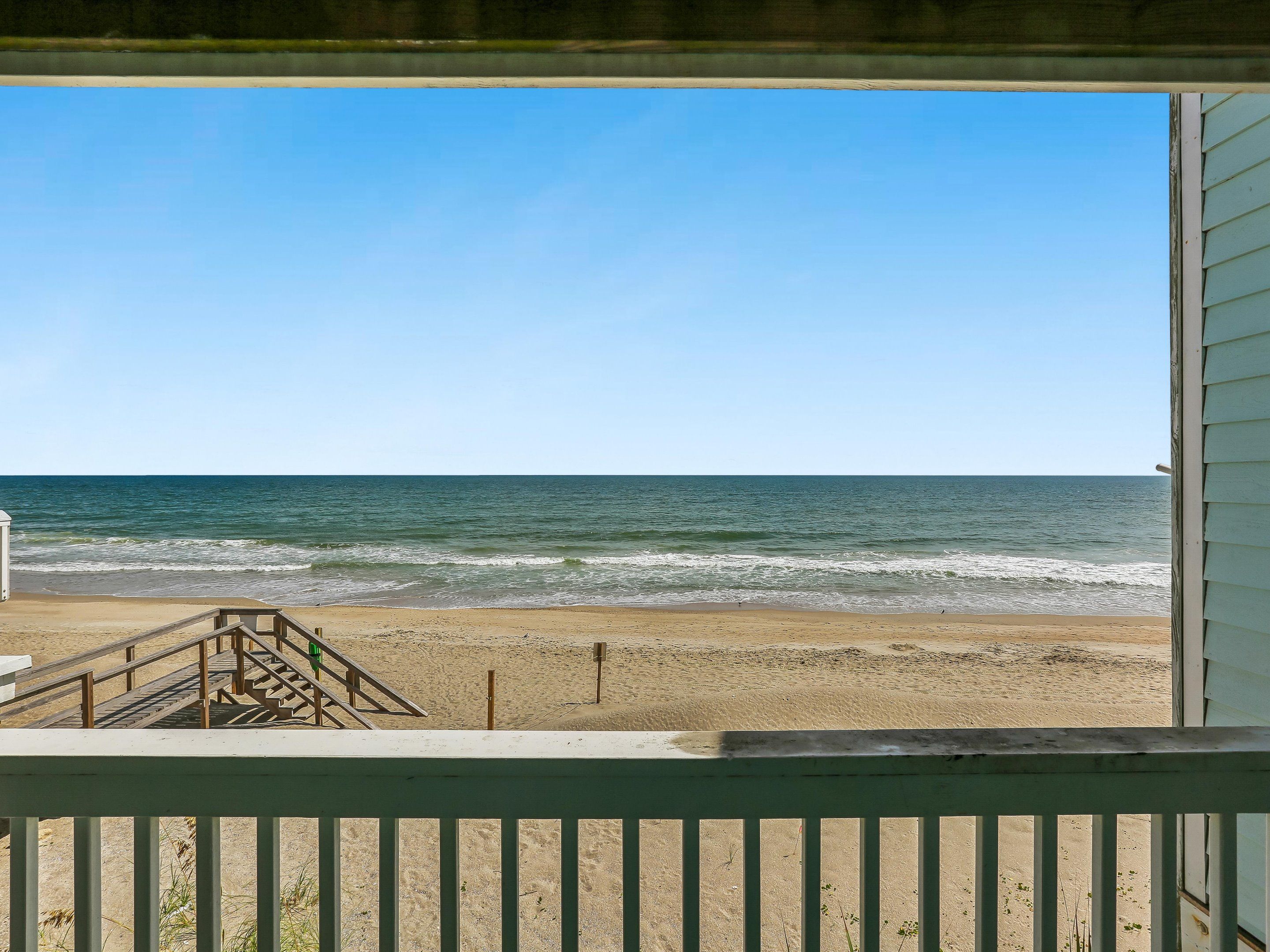 Vista da praia a partir de uma varanda com guarda-corpo de madeira, areia dourada e o oceano.