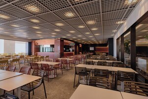 Uma ampla área de refeições interna com várias mesas e cadeiras, um design de teto moderno e grandes janelas que proporcionam iluminação natural.