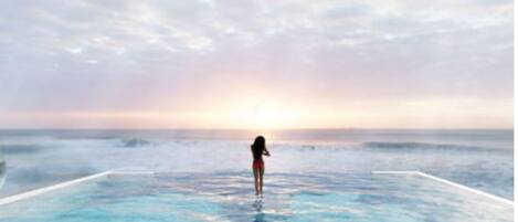 Uma pessoa em uma piscina infinita com fundo de vidro, com vista para o oceano e um pôr do sol ao fundo.