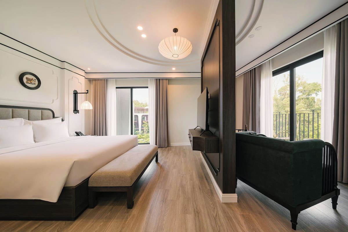 Quarto de hotel com uma cama grande, um sofá, uma TV e uma varanda com cortinas.