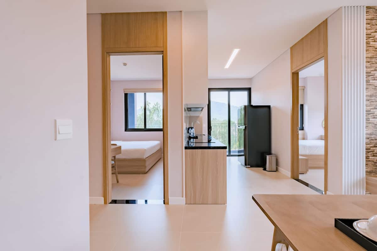 Quarto de hotel moderno com uma mesa de jantar de madeira, uma área de cozinha e um quarto visível por uma porta aberta.