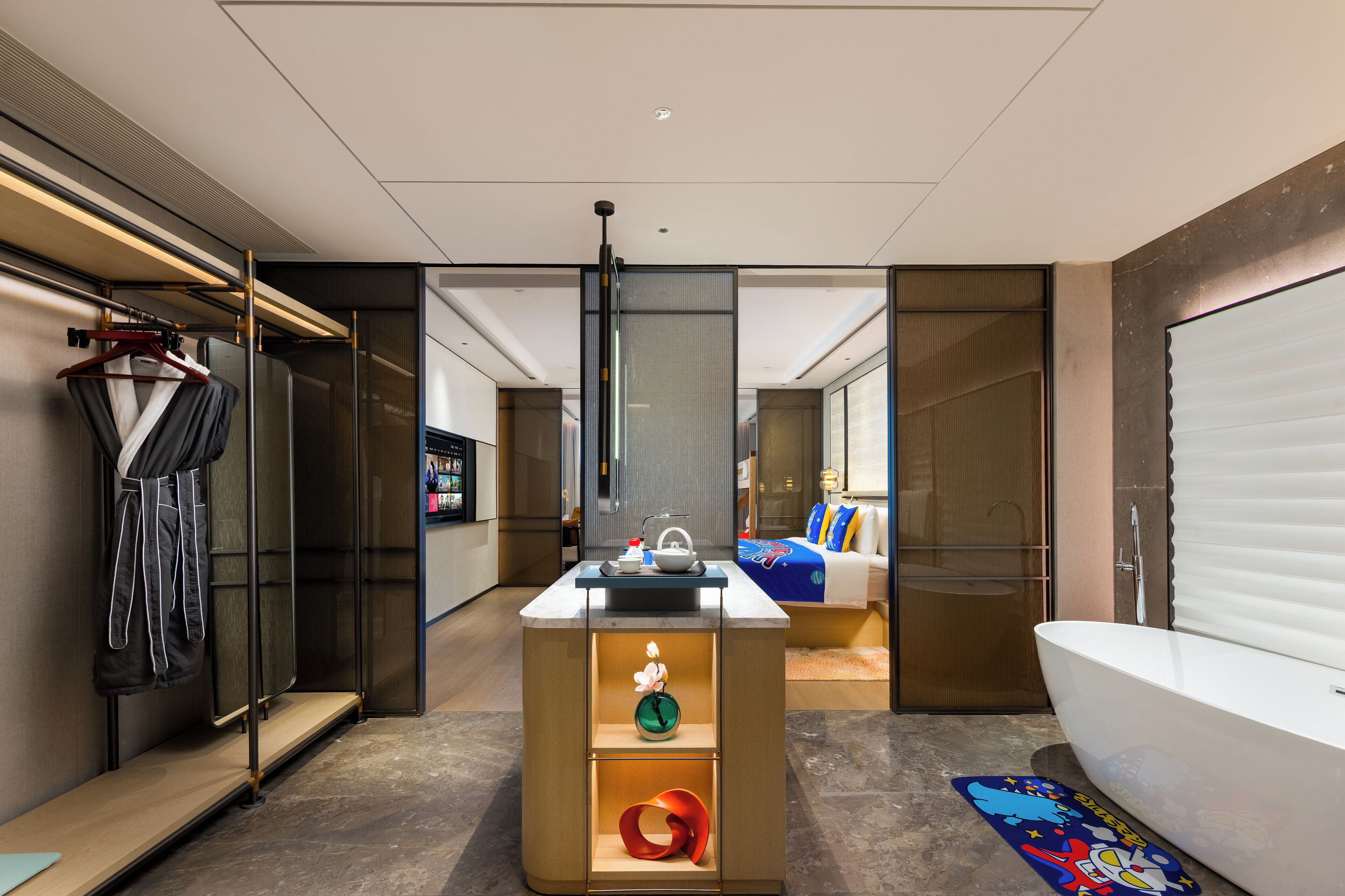 Quarto de hotel moderno com uma banheira grande, uma secretária pequena com uma lâmpada, um guarda-roupa e uma cama com colcha azul e branca.