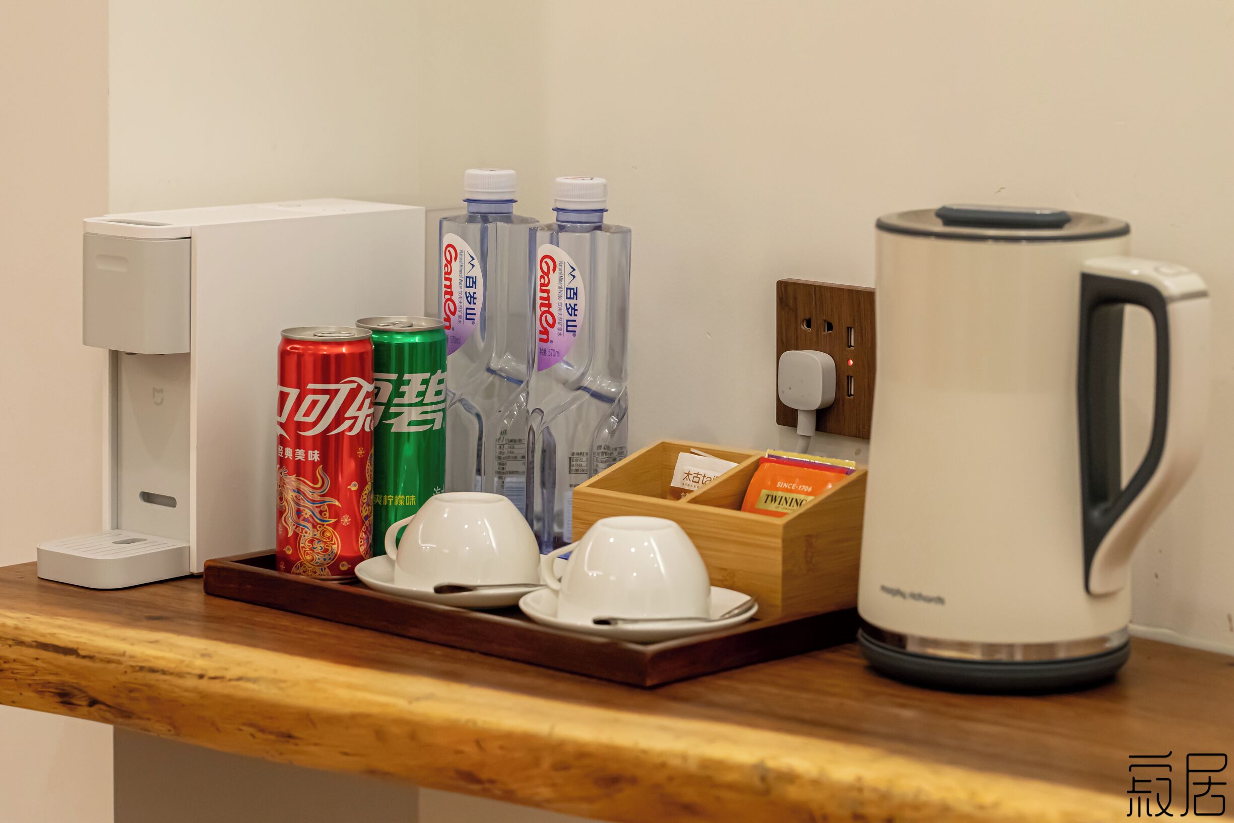 Espaço de café da manhã em quarto de hotel, com cafeteira, duas latas de refrigerante, duas garrafas de água, chaleira e bandeja com xícaras e pires.