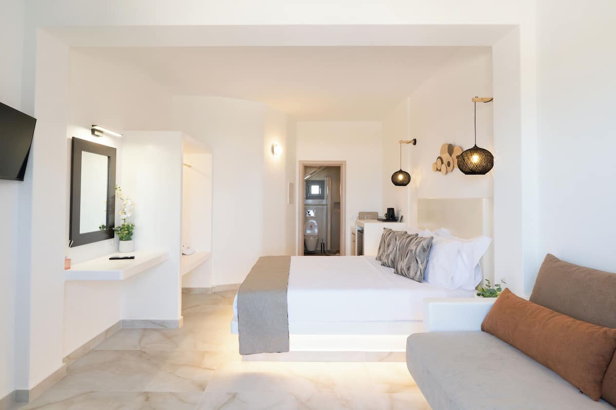 Quarto moderno com cama grande, sofá, televisão e banheiro visível ao fundo.