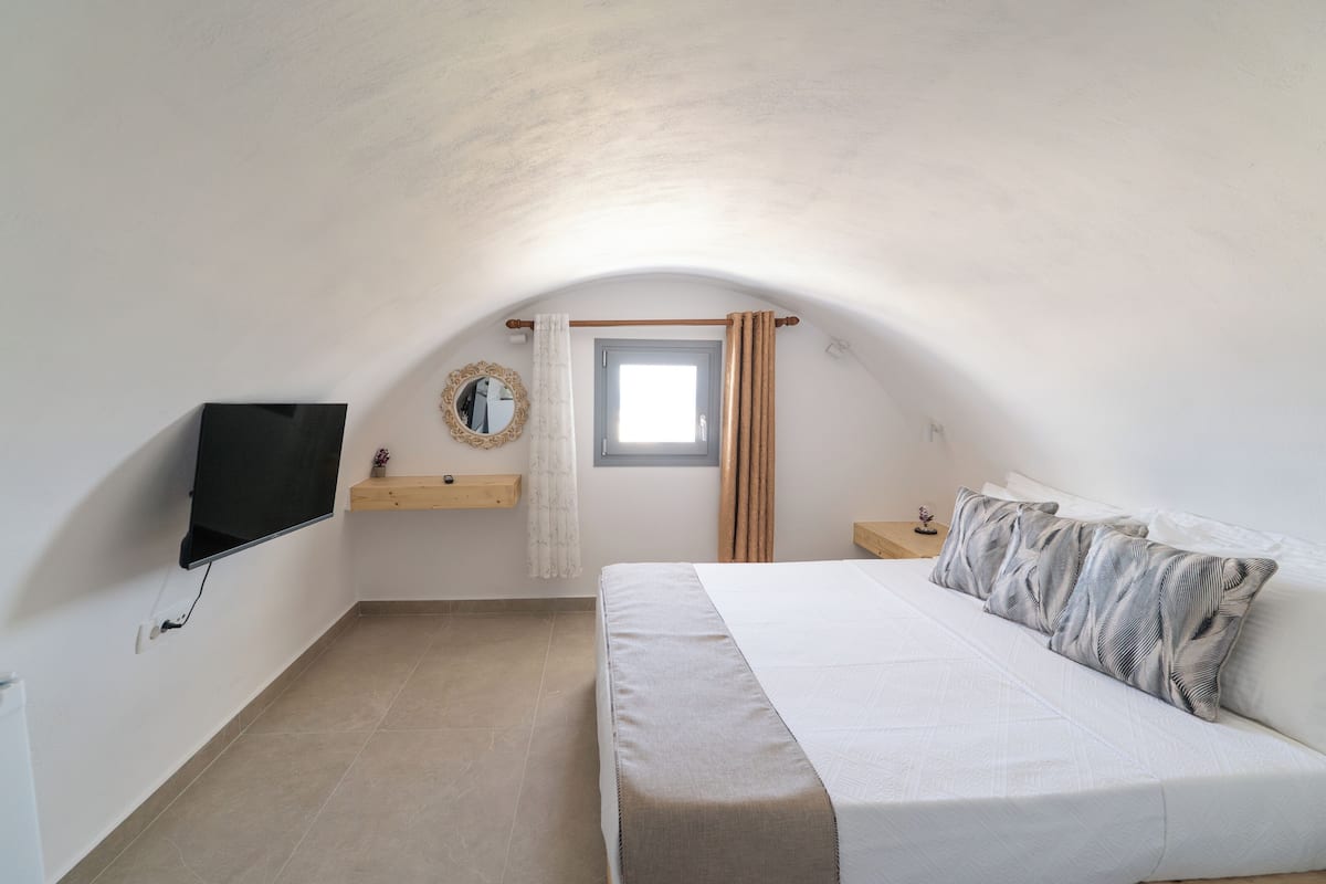 Quarto com cama, televisão fixada na parede, janela com cortinas e uma pequena estante com espelho e planta.