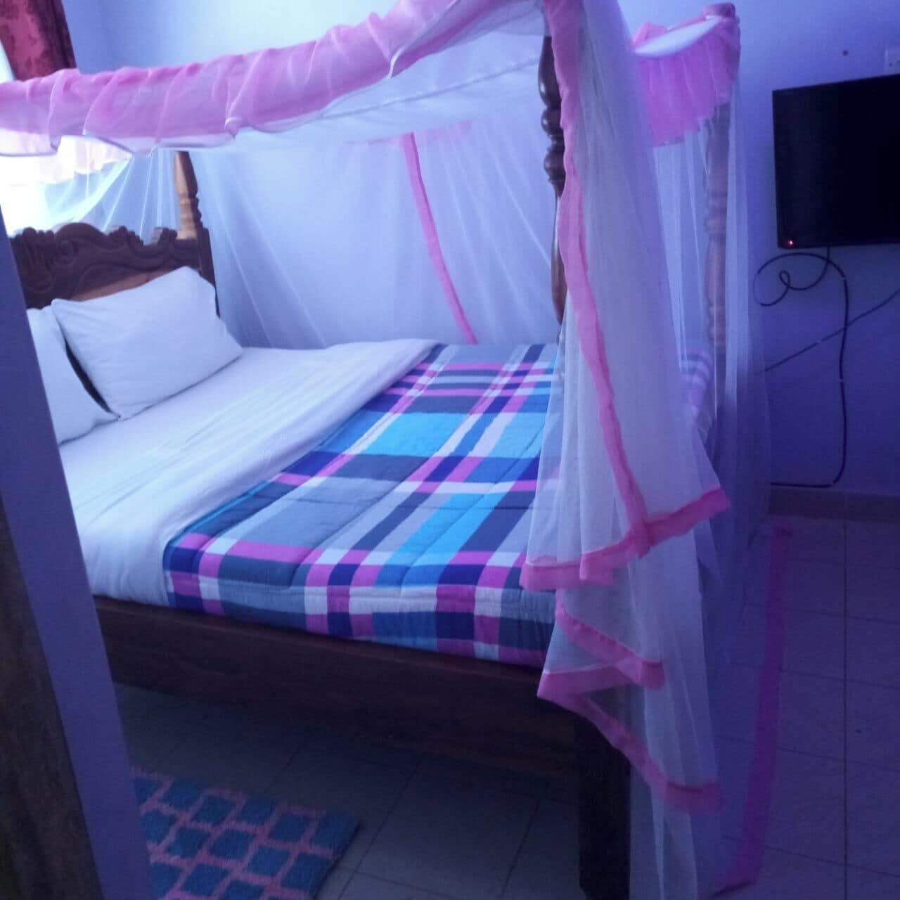 Uma cama com colcha xadrez, dossel branco e televisão na parede.