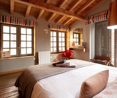 Quarto com teto de madeira, janelas amplas com cortinas listradas, uma cadeira vermelha e cama com colcha branca e castanha.