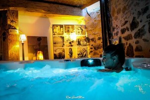 Uma pessoa em uma banheira de hidromassagem, com uma parede de pedra e uma prateleira com diversos objetos ao fundo.