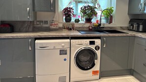 Uma cozinha moderna com armários cinzentos, máquina de lavar embutida e uma janela com cortina florida.