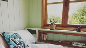 Cama com lençóis brancos, bandeja com uma chávena e um saquinho de chá, janela com vista para vegetação, estante com livros e um pássaro decorativo.