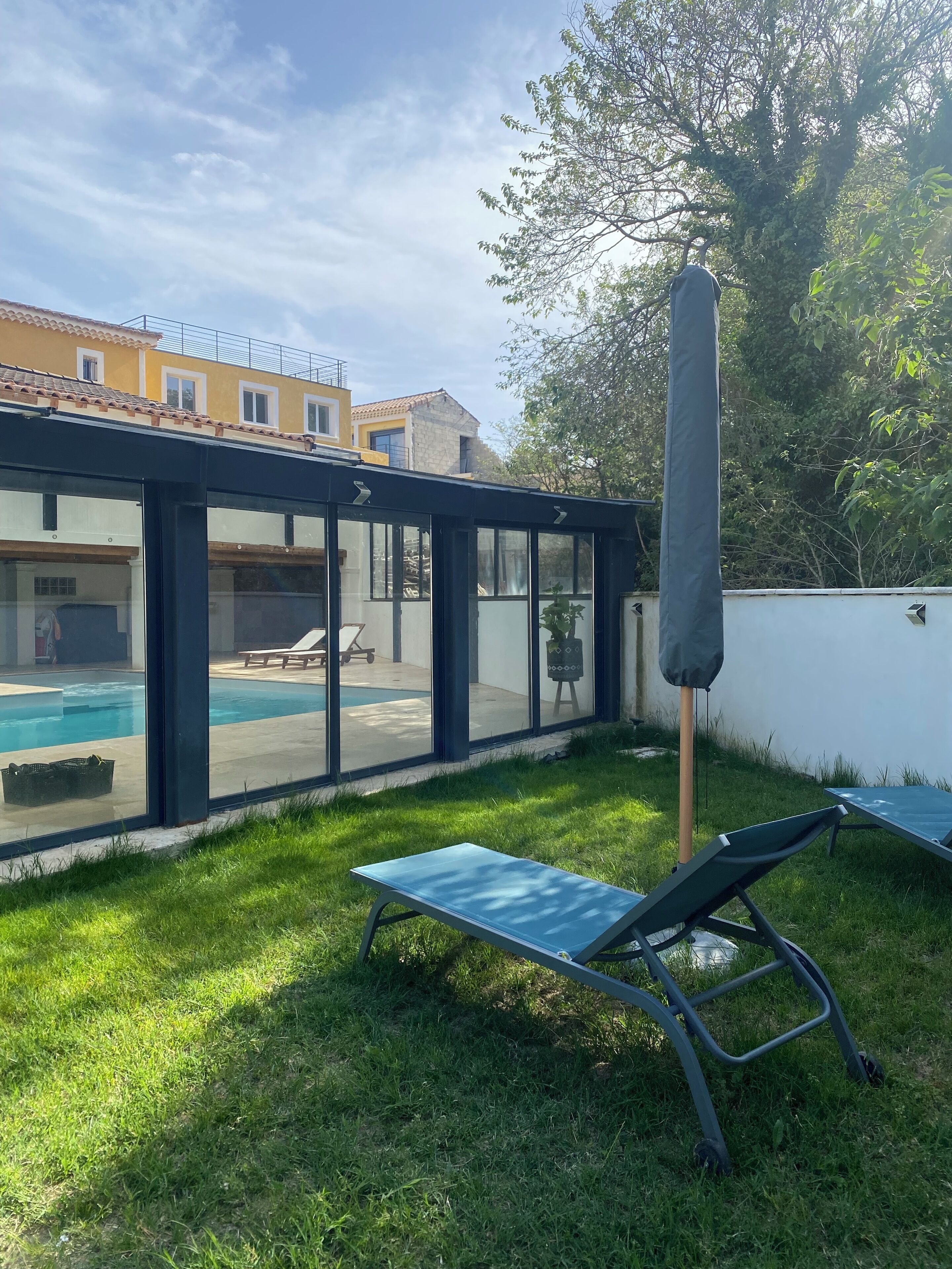 Uma área à beira da piscina com uma estrutura de vidro, uma espreguiçadeira e uma mesa coberta.