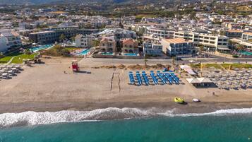 Resort costeiro com vários edifícios, uma praia com guarda-sóis e vista clara para o mar.