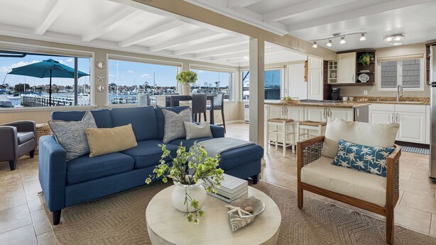 Uma sala de estar ampla com um sofá azul, uma cadeira de madeira e uma mesa de centro com objetos decorativos. Uma grande janela oferece vista para um porto de iates.