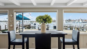Uma sala de jantar com uma mesa e cadeiras, um vaso com flores e vista para uma marina com barcos e uma ponte.