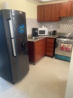 Uma cozinha com geladeira de aço inoxidável, micro-ondas, fogão a gás, armários de madeira e painel de azulejos.