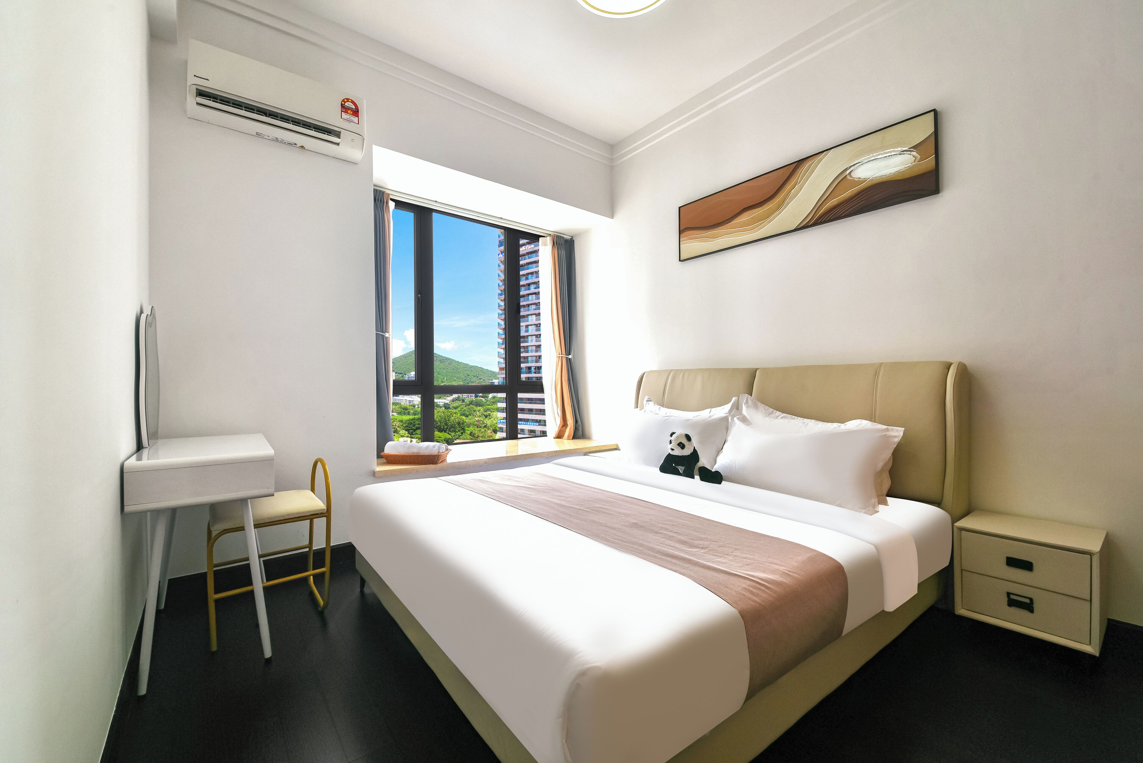 Quarto de hotel com cama, escrivaninha, cadeira, mesa de cabeceira e janela com vista para edifícios e vegetação.