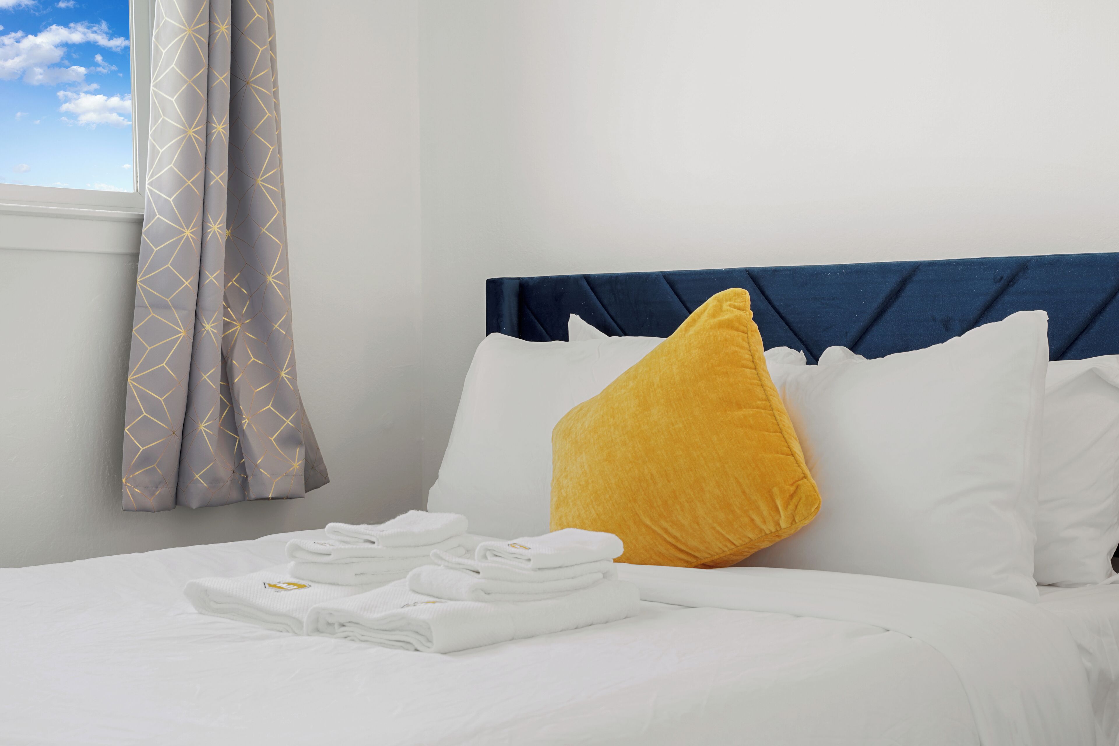 Cama bem arrumada com lençóis brancos e toalha dobrada, cabeceira azul e travesseiro amarelo.
