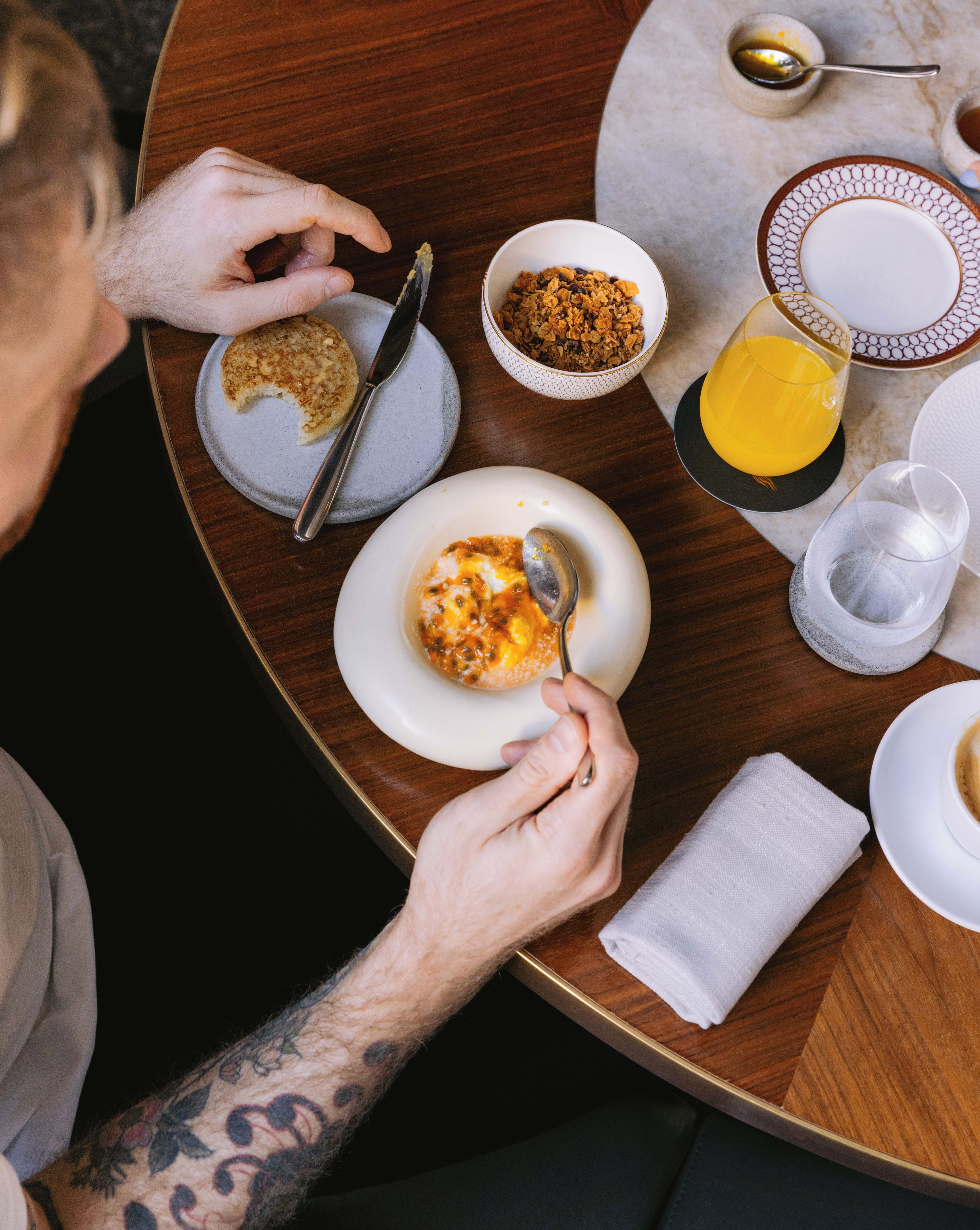 Uma pessoa saboreando o café da manhã em uma mesa redonda, com um prato de comida, tigela de cereal, copo de suco de laranja e uma colher.