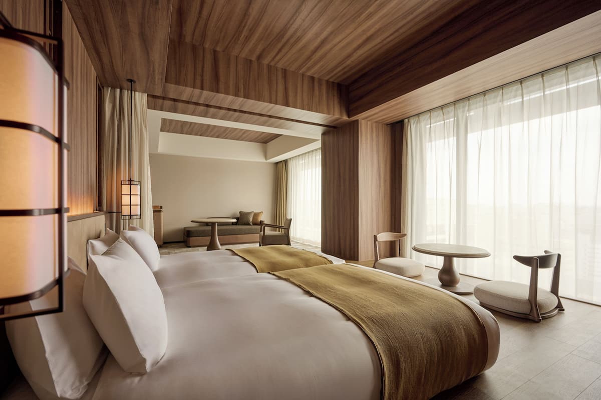 Quarto de hotel moderno com uma cama grande, um espaço de estar com sofá e uma mesa pequena, e vista para a cidade através de janelas amplas.