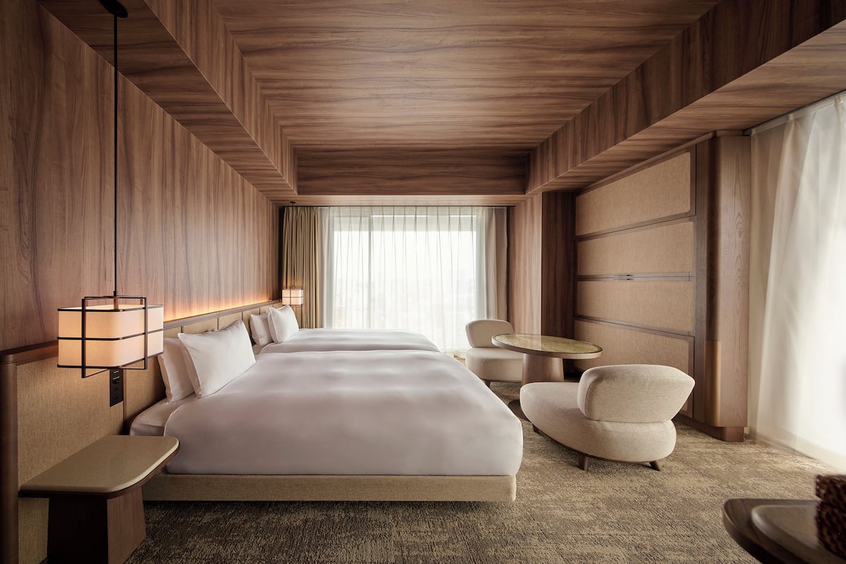 Quarto de hotel com uma cama grande, uma mesinha de cabeceira, uma cadeira, uma mesa redonda pequena e uma janela com cortinas.