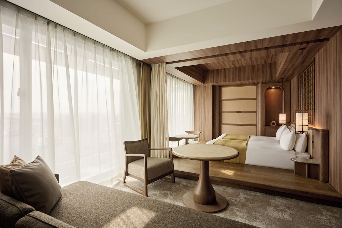 Quarto de hotel moderno com sofá, mesa redonda, cama com almofadas e parede de destaque em madeira.