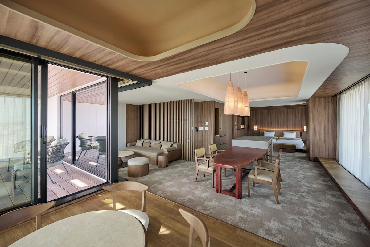 Quarto de hotel moderno com uma grande porta de vidro deslizante que dá para um terraço, área de estar aconchegante com sofá e poltronas, mesa de jantar com cadeiras, e um quarto separado com cama e mesas de cabeceira.