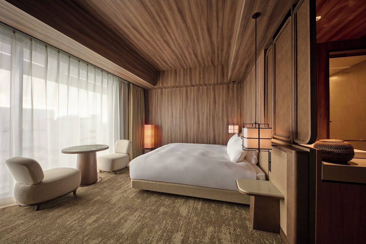 Quarto de hotel moderno com uma cama grande, duas poltronas, uma mesinha e uma janela ampla com cortinas leves.