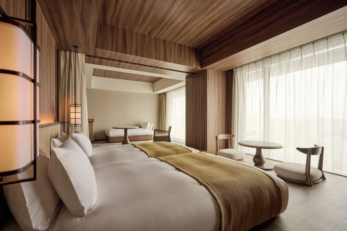 Quarto de hotel moderno com uma cama grande, área de estar com mesa e cadeiras, e janelas amplas com cortinas leves.