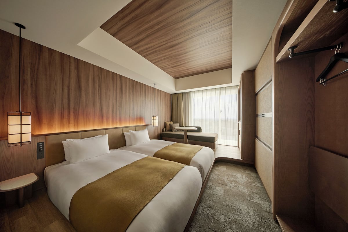 Quarto de hotel com duas camas, uma mesinha, um sofá e uma janela grande com cortinas.