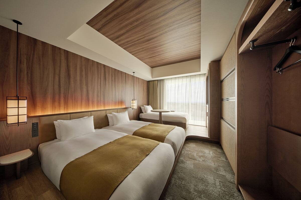 Quarto de hotel com duas camas, uma mesa pequena, um sofá, um lustre e uma janela grande com cortinas.