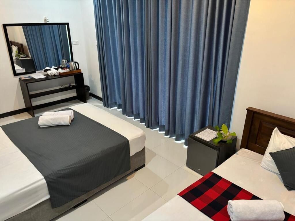 Quarto de hotel com duas camas, uma escrivaninha com espelho, uma mesinha de centro com planta e cortinas azuis.