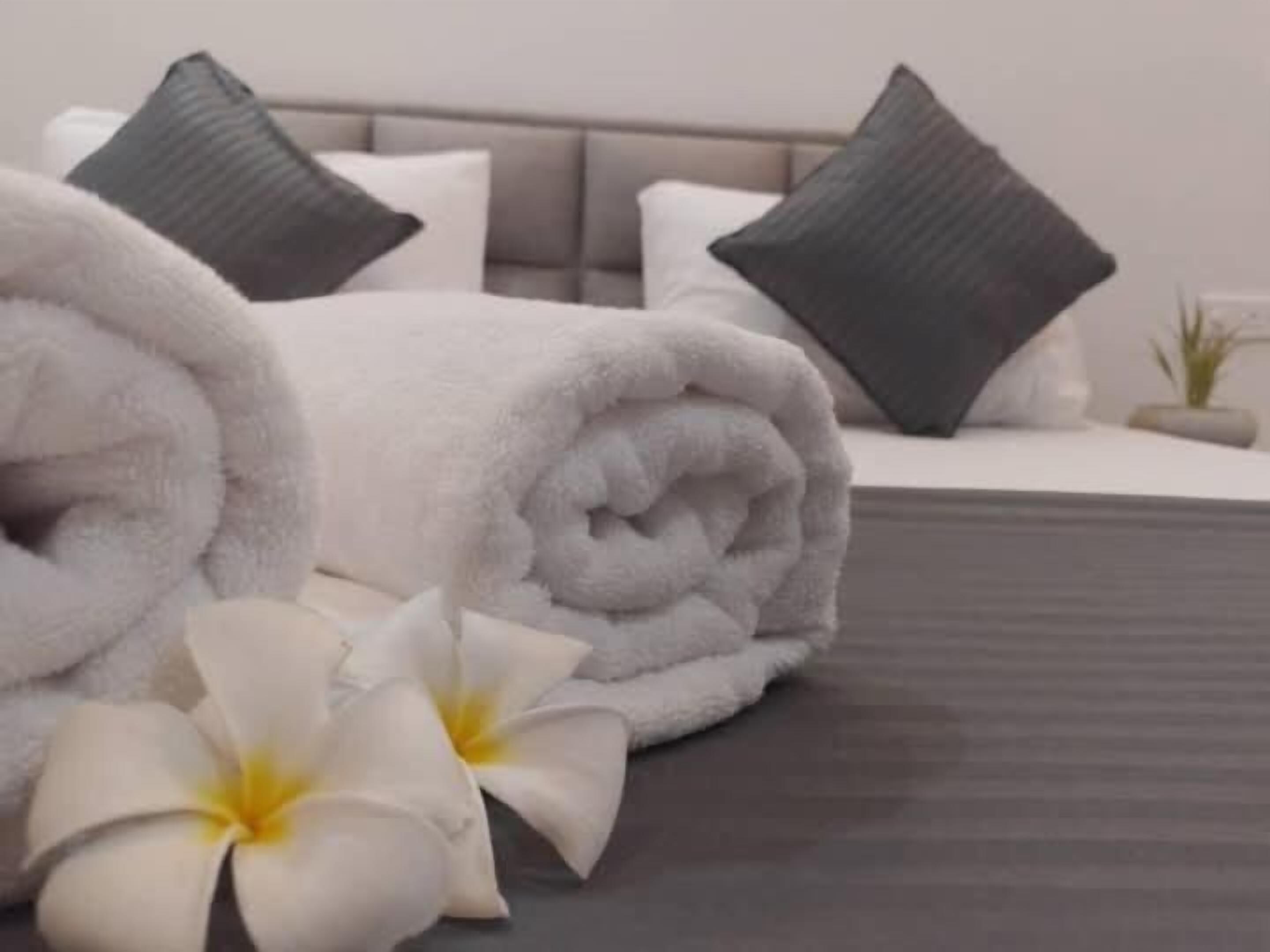 Cama arrumada com toalhas brancas dobradas, almofadas escuras, uma pequena planta em vaso e flores brancas sobre a cama.